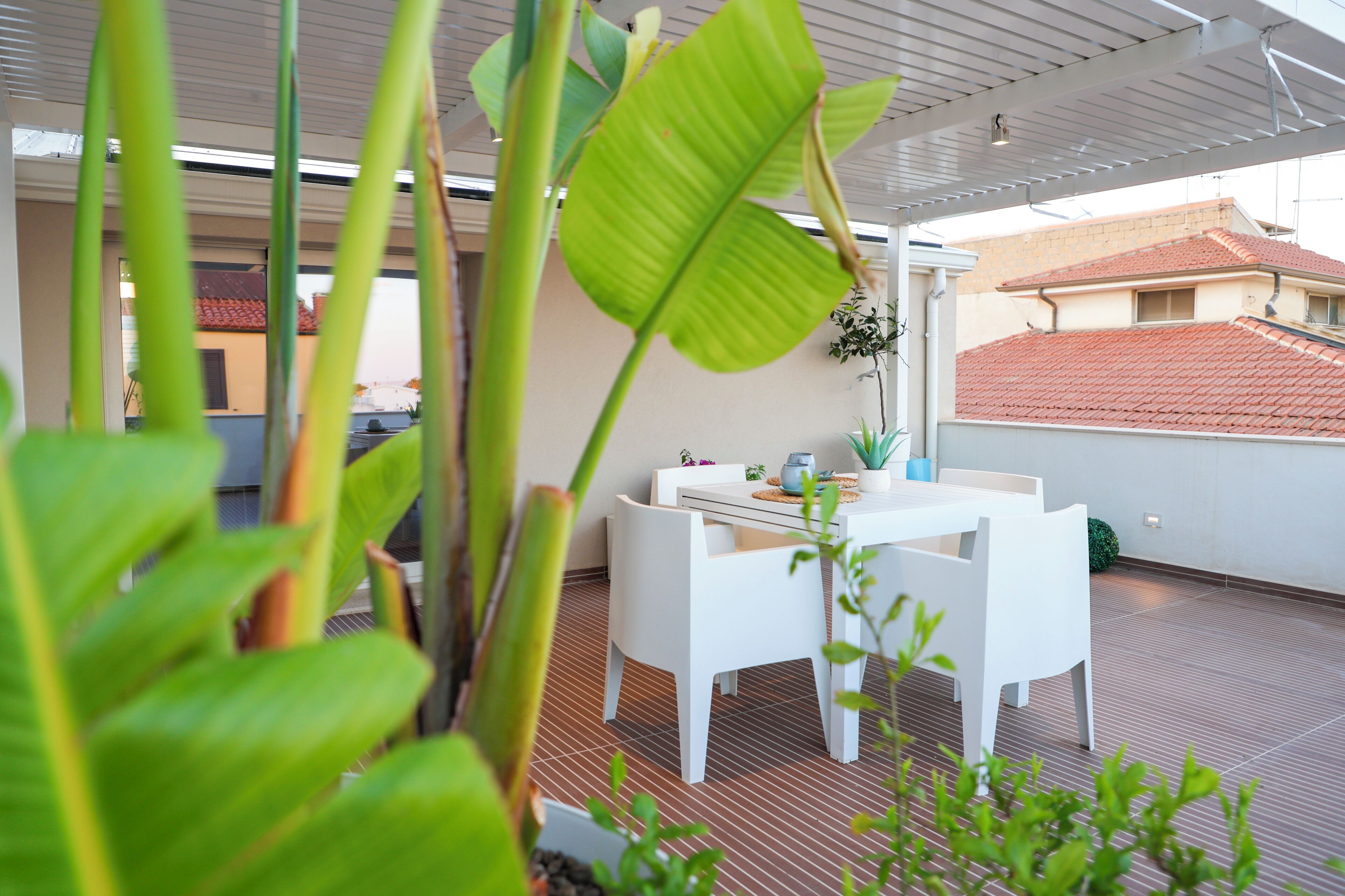 Terraço com mesa e cadeiras brancas, rodeado por plantas em vasos.