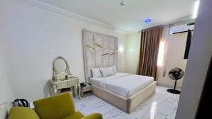 Quarto de hotel com uma cama, uma mesinha de toucador com espelho, uma poltrona amarela, um ventilador de chão e piso de mármore.