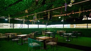 Uma área de refeições interna com mesas e cadeiras, piso de grama sintética verde e plantas penduradas.