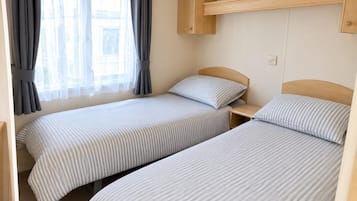 Quarto com duas camas de solteiro, cabeceira de madeira, prateleira pequena e janela com cortinas.