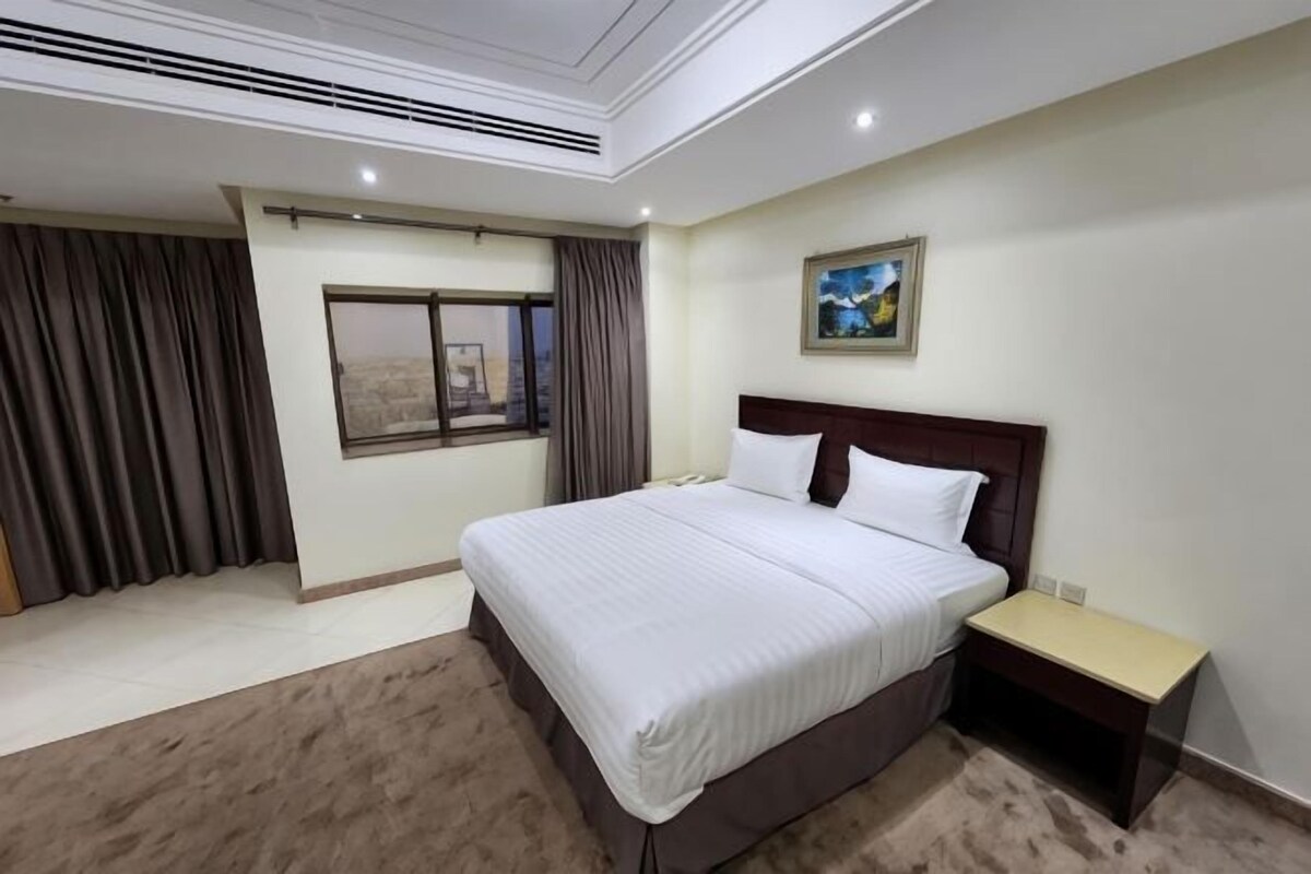 Quarto de hotel com uma cama grande, mesa de cabeceira, janela com cortinas e um quadro na parede.