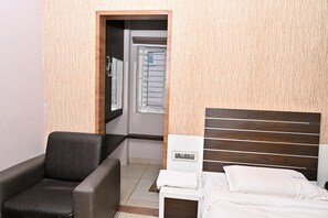 Quarto de hotel com cama, uma cadeira, janela com cortinas e porta que dá para outro cômodo.