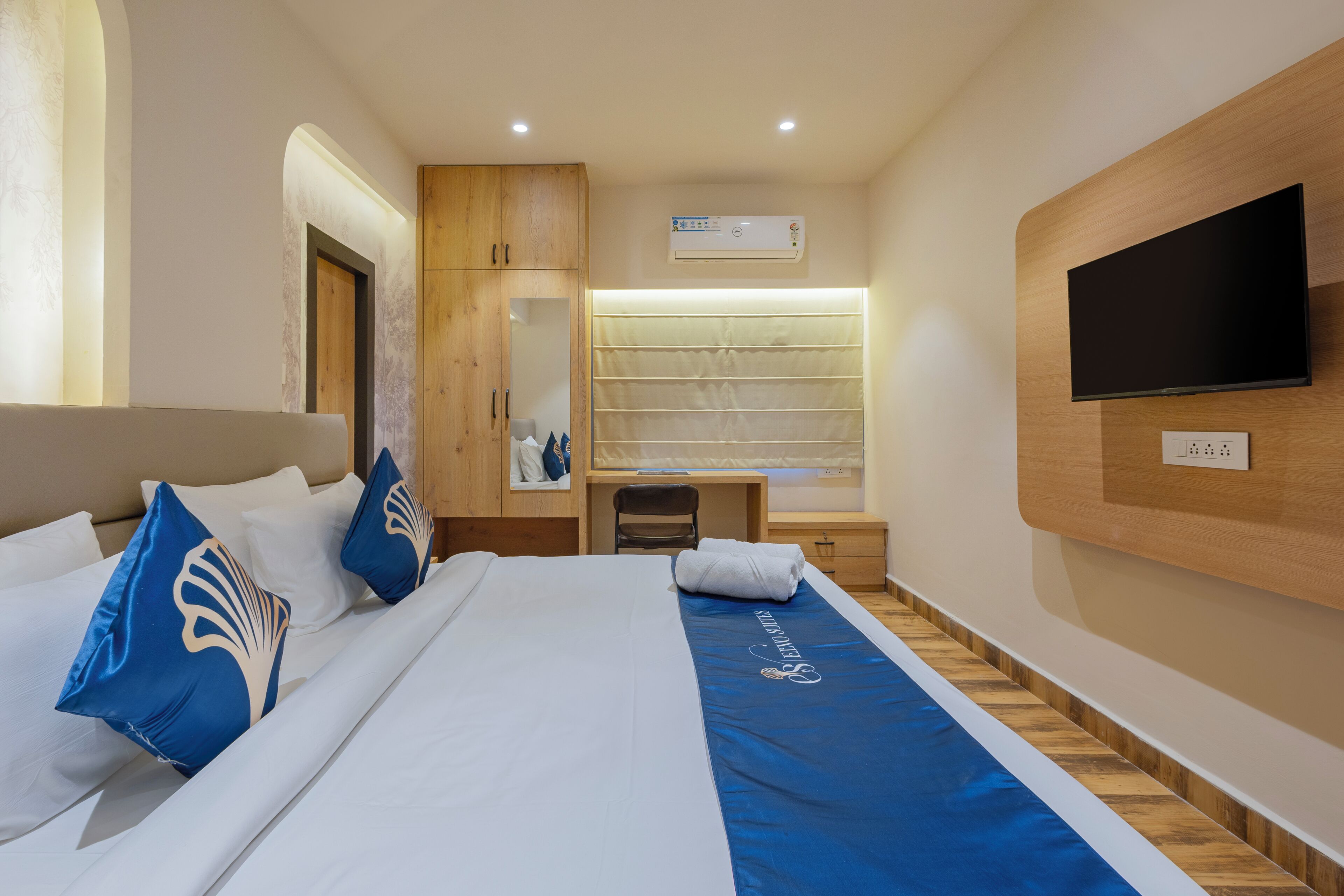 Quarto de hotel com cama, escrivaninha, cadeira, televisão fixada na parede e janela com persianas.