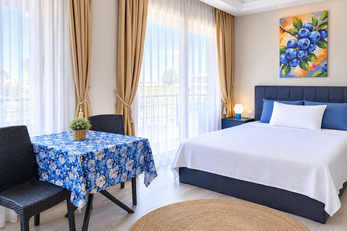 Quarto de hotel com cama, mesa com toalha de mesa florida, cadeira, pintura de mirtilos e vista para o exterior.