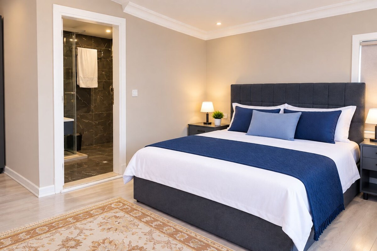Quarto com cama, mesas de cabeceira, cabeceira escura, banheiro com chuveiro de vidro e um guarda-roupa cinzento.