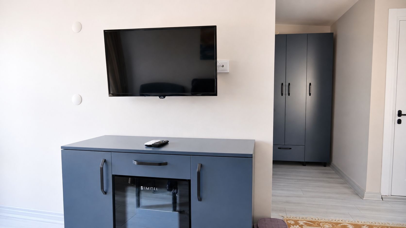 Quarto com televisão de tela plana na parede, um armário cinza com dispensador de água e um guarda-roupa com duas portas.
