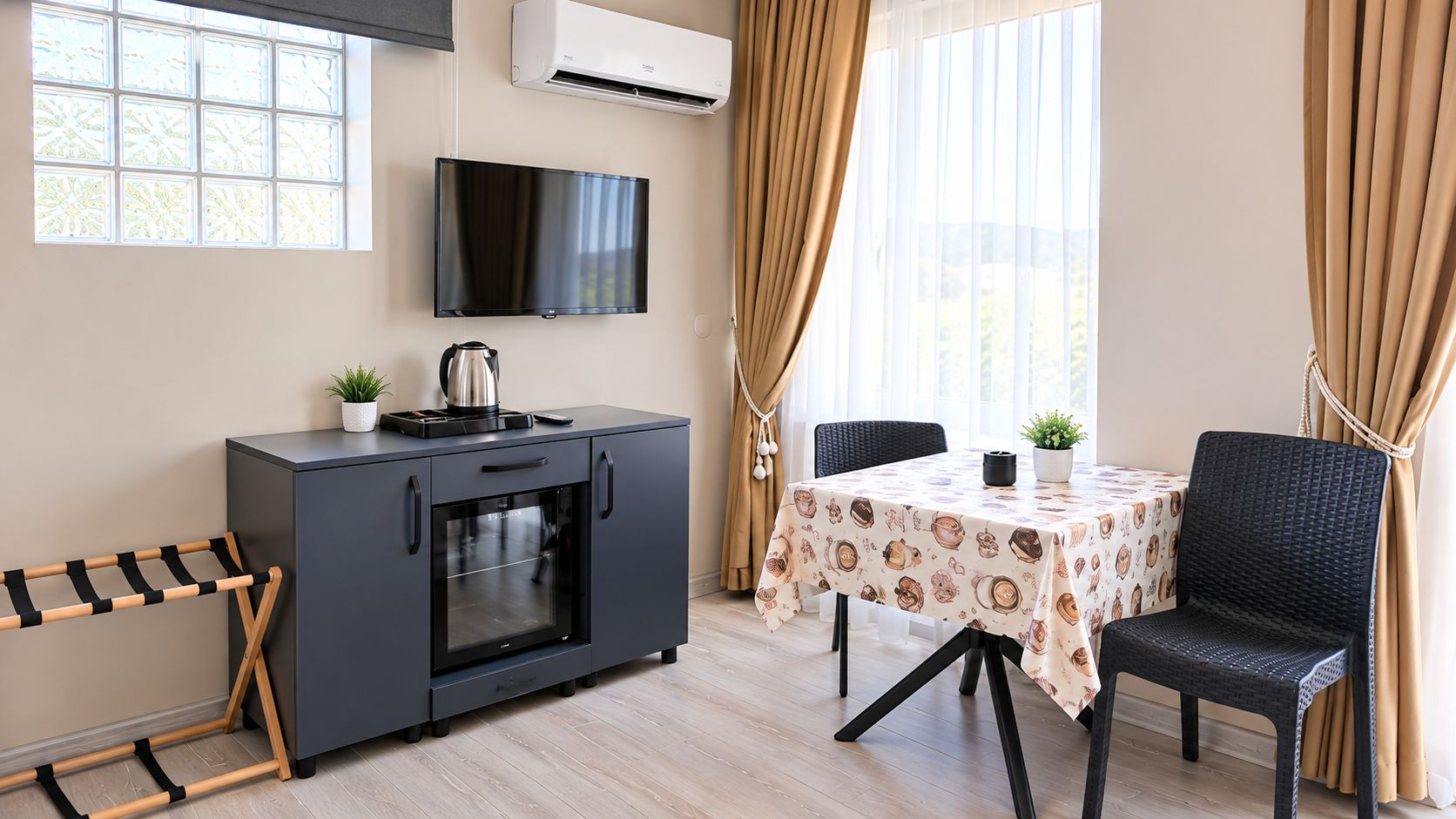 Cozinha compacta com forno embutido, máquina de café e televisão acima. Há uma mesa de jantar com cadeiras e uma janela com cortinas.