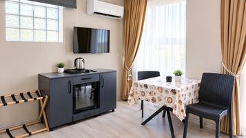 Cozinha compacta com forno embutido, máquina de café e televisão acima. Há uma mesa de jantar com cadeiras e uma janela com cortinas.