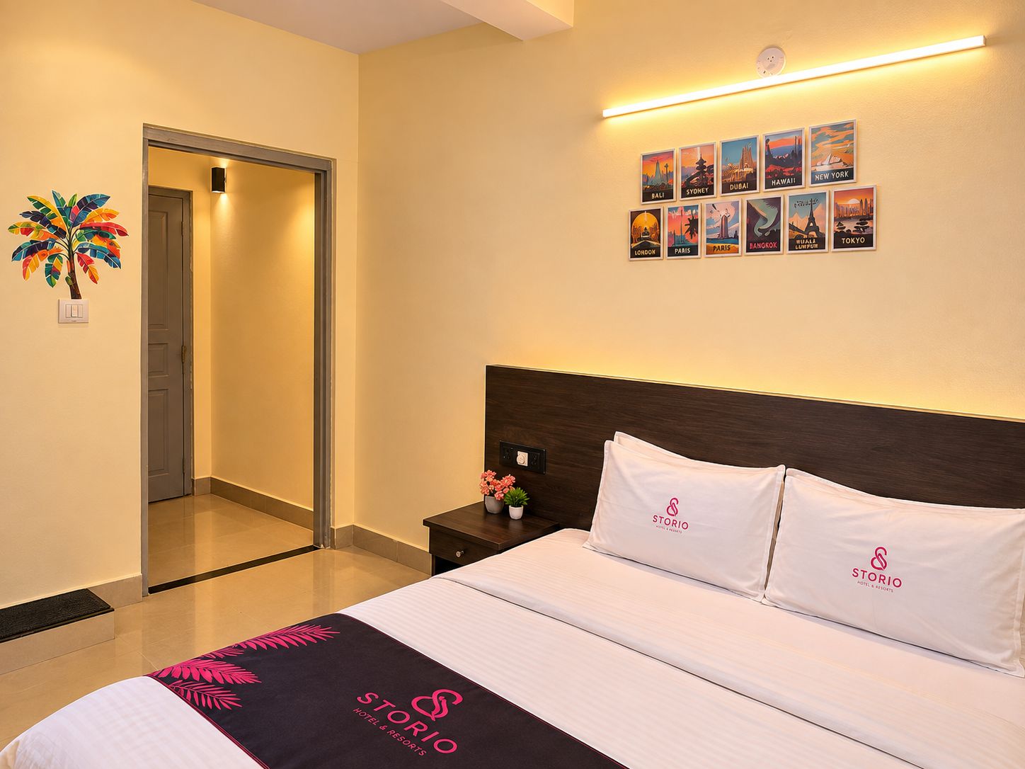 Quarto de hotel com cama, mesa de cabeceira, relógio na parede e uma coleção de cartazes de viagem.