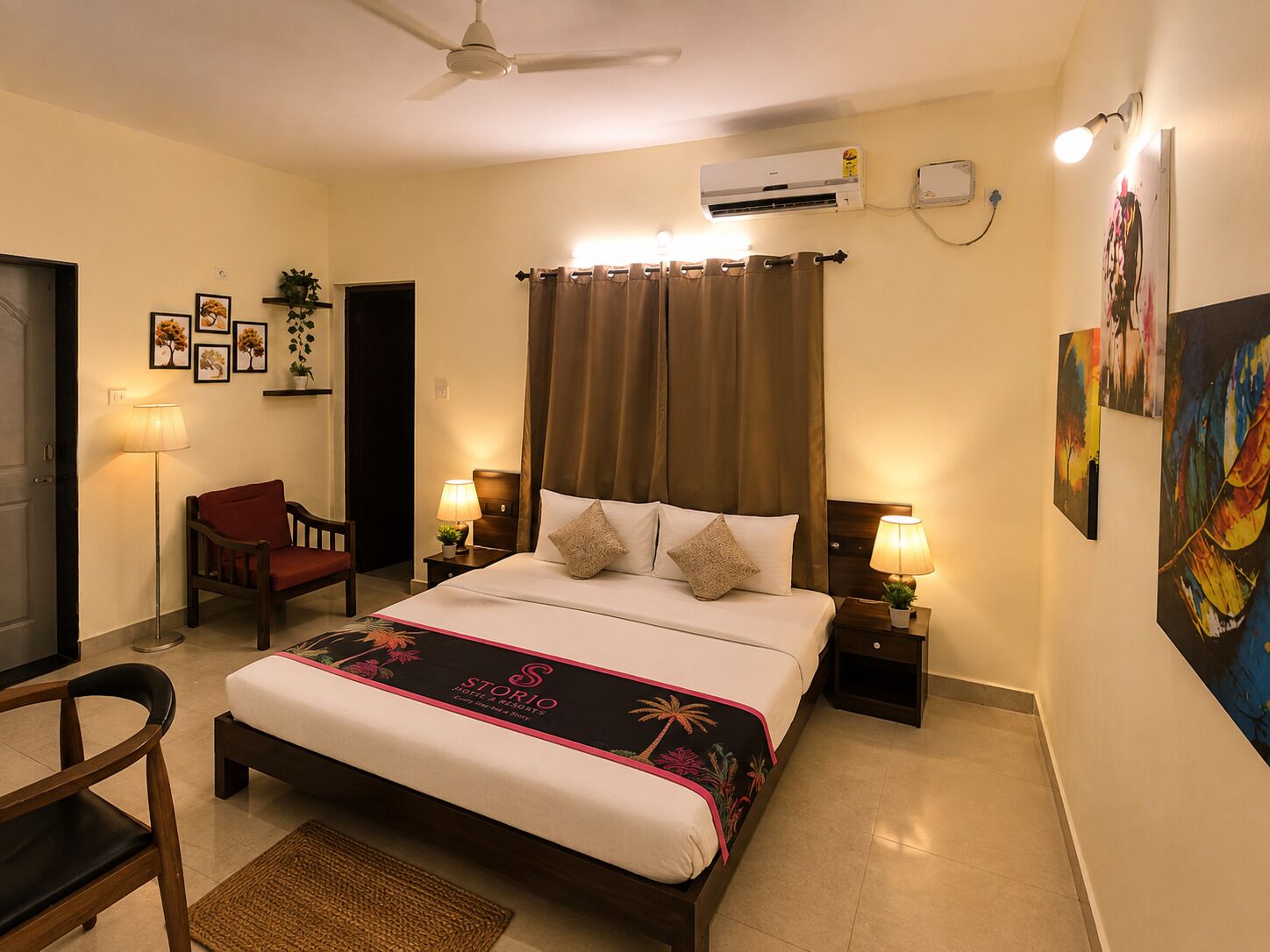 Quarto de hotel com uma cama, duas mesas de cabeceira, uma cadeira, ar condicionado e obras de arte nas paredes.