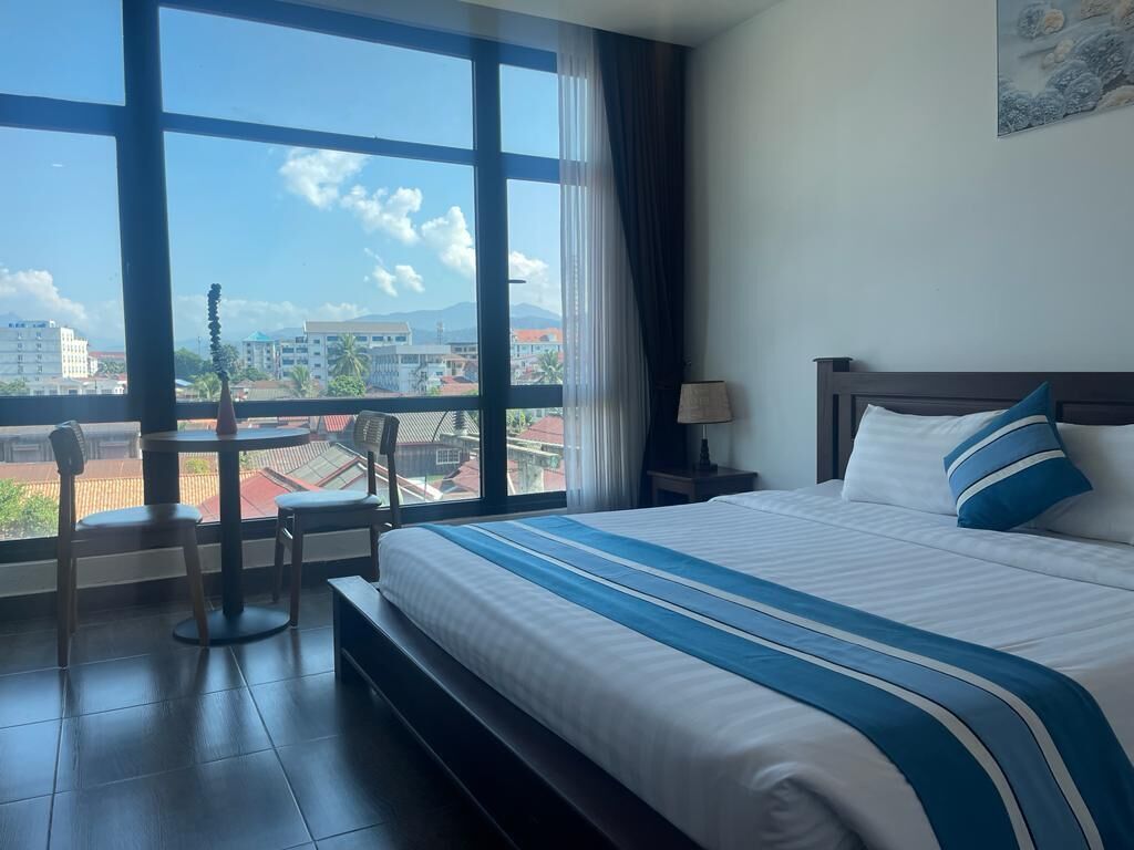 Quarto de hotel com uma janela grande, cama com roupa de cama listrada, mesa de cabeceira e uma cadeira.