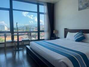 Quarto de hotel com uma janela grande, cama com roupa de cama listrada, mesa de cabeceira e uma cadeira.