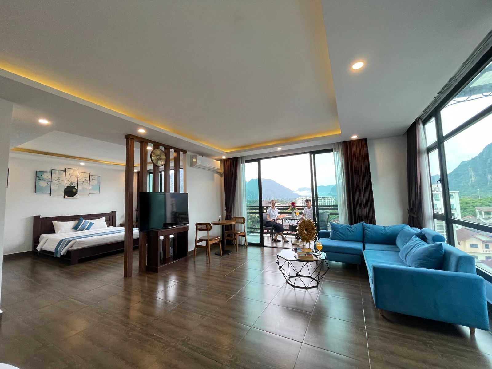 Quarto de hotel moderno com cama, TV, sofá, mesa de jantar com cadeiras e uma grande janela com vista para as montanhas.