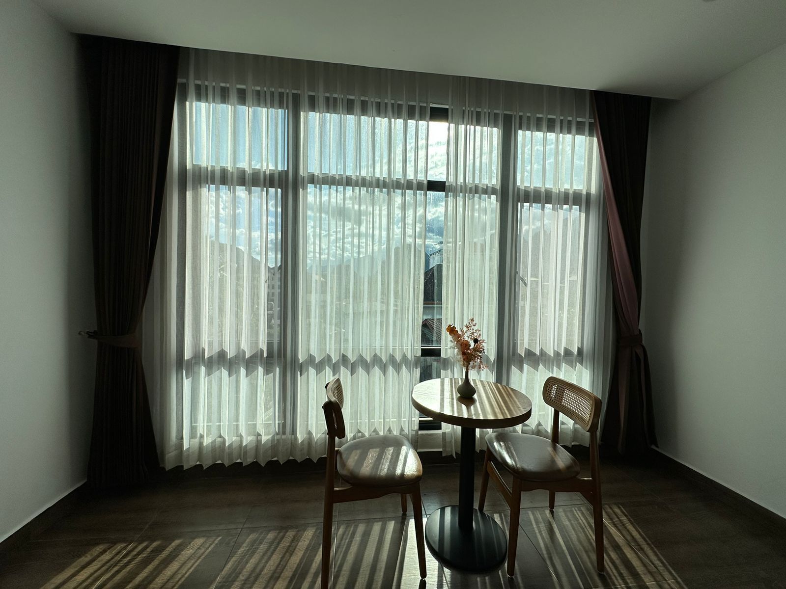 Quarto com mesa redonda e duas cadeiras, vaso com flores e vista através de amplas janelas com cortinas leves.
