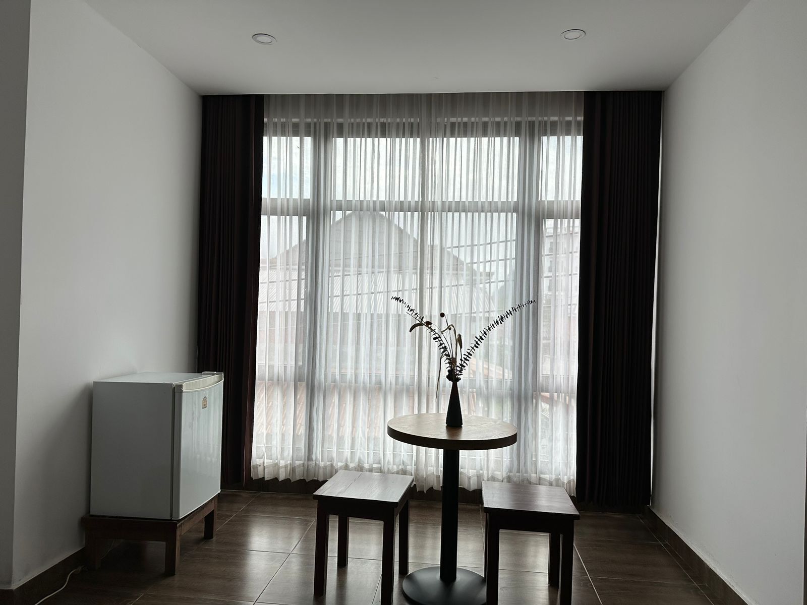 Quarto com mesa de jantar e cadeiras, um pequeno armário e uma janela grande com cortinas leves.