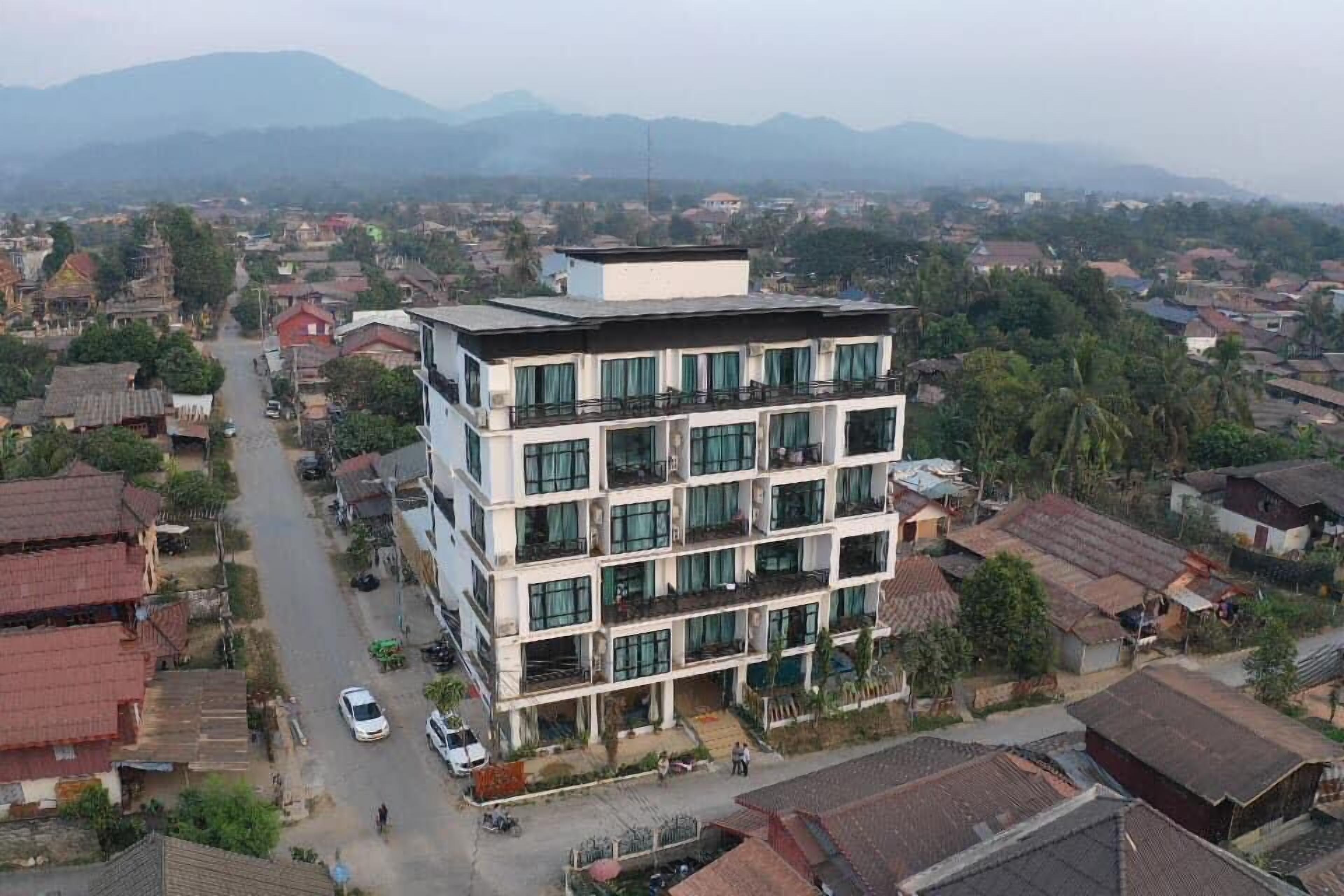Um edifício com vários andares e varandas, rodeado por casas residenciais e vegetação.
