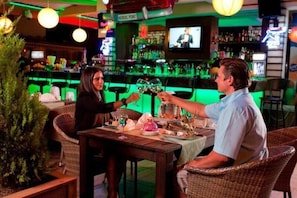 Uma área de bar com uma tela de televisão, prateleiras repletas de garrafas e cadeiras de vime ao redor de uma mesa de madeira, com iluminação verde ao fundo.