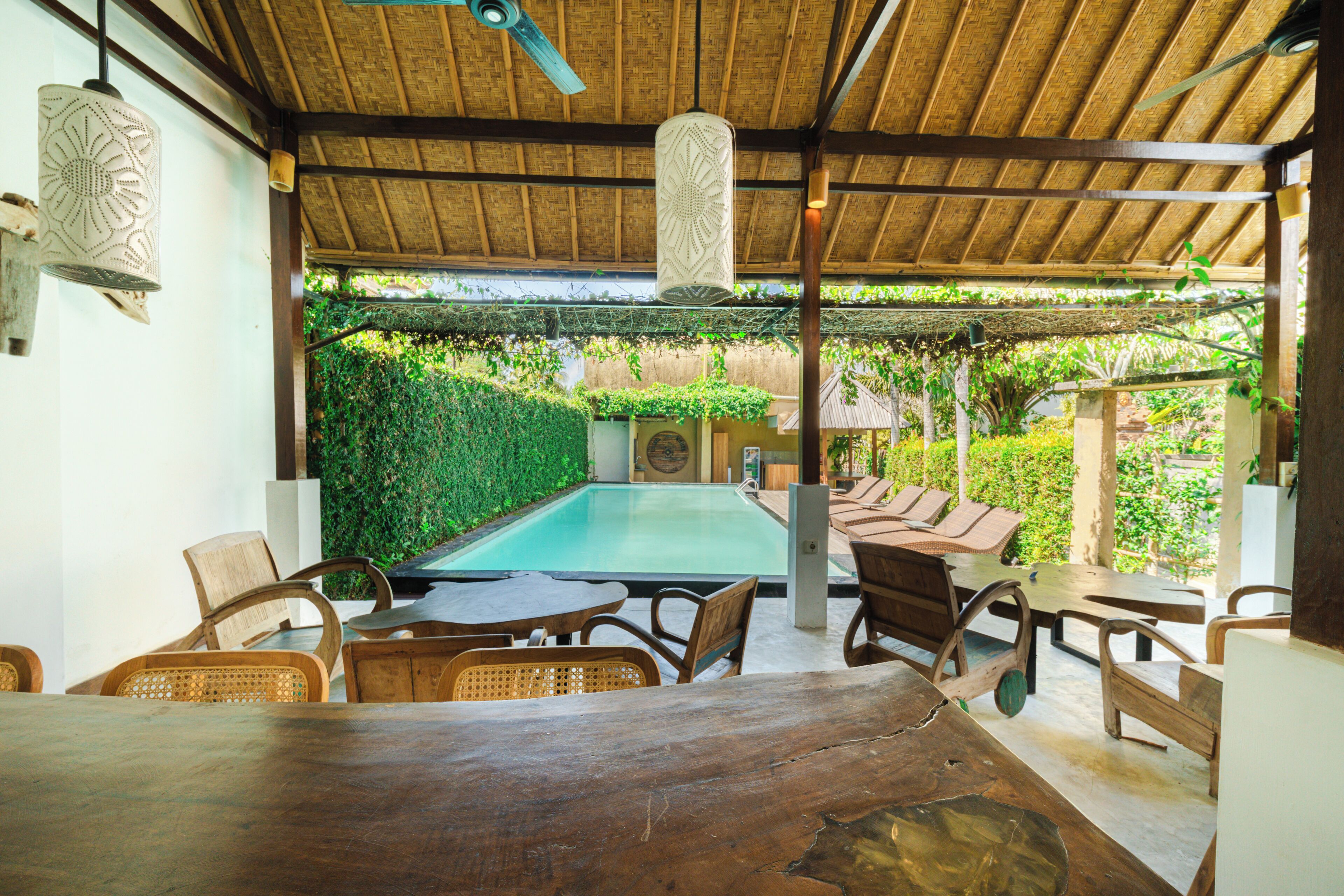 Área de piscina ao ar livre com um telhado de palha, cercada por vegetação exuberante e mobiliada com cadeiras e mesas de madeira.