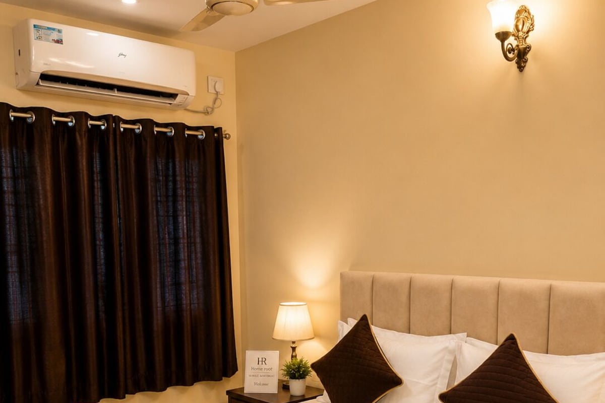 Quarto de hotel com cama, mesa de cabeceira, luminária, iluminação embutida na parede, ar condicionado e ventilador de teto.