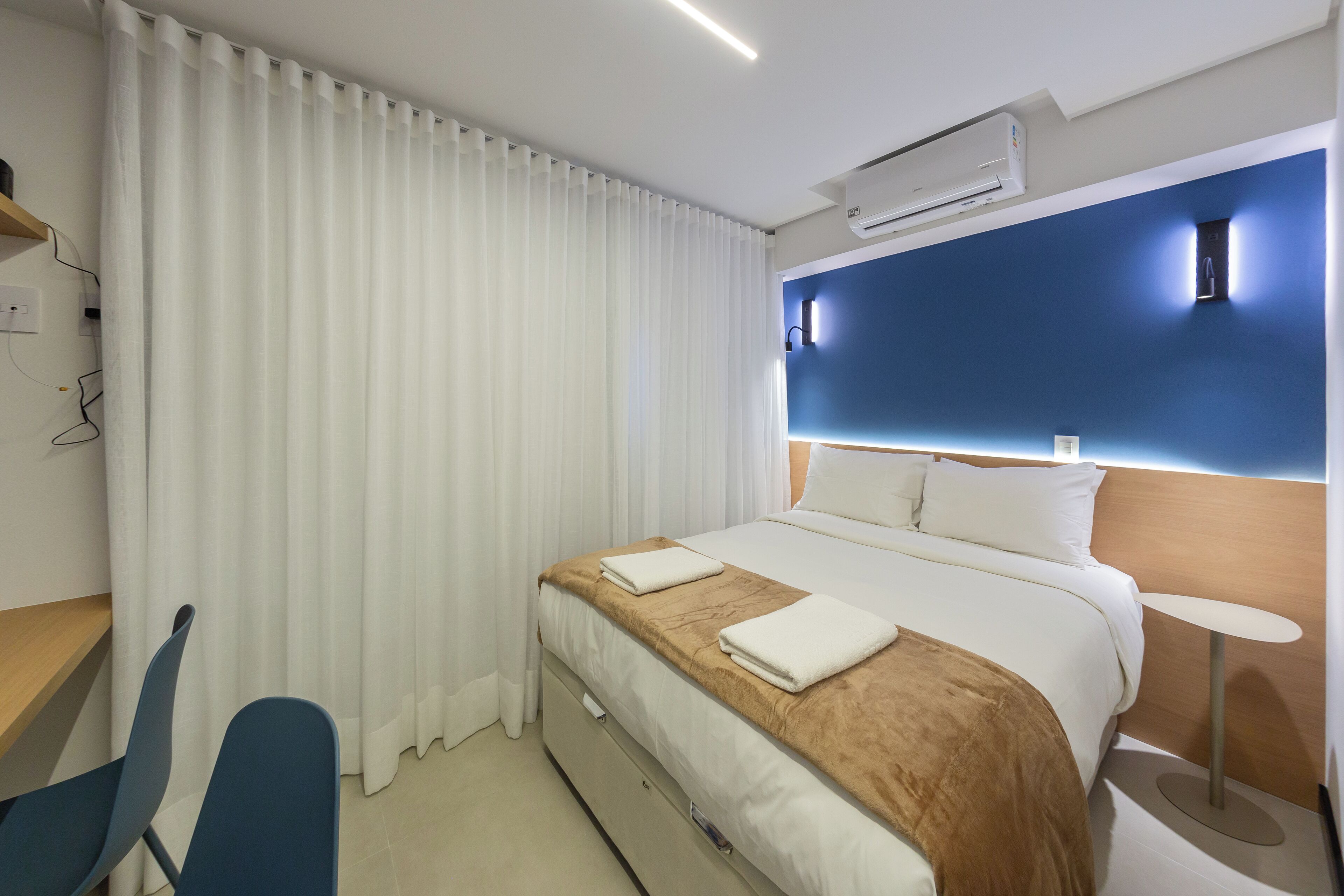 Quarto de hotel com cama, escrivaninha, cadeira, ar condicionado e luminária na parede.