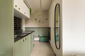 Um corredor estreito com uma área de cozinha de um lado, um banco verde e um espelho grande na parede oposta.