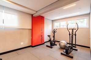 Uma academia com equipamentos de ginástica, uma parede vermelha com texto motivacional e uma janela com vista.
