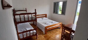 Quarto com duas camas beliche de madeira, uma cama de solteiro, uma mesa de madeira e uma janela com persianas.