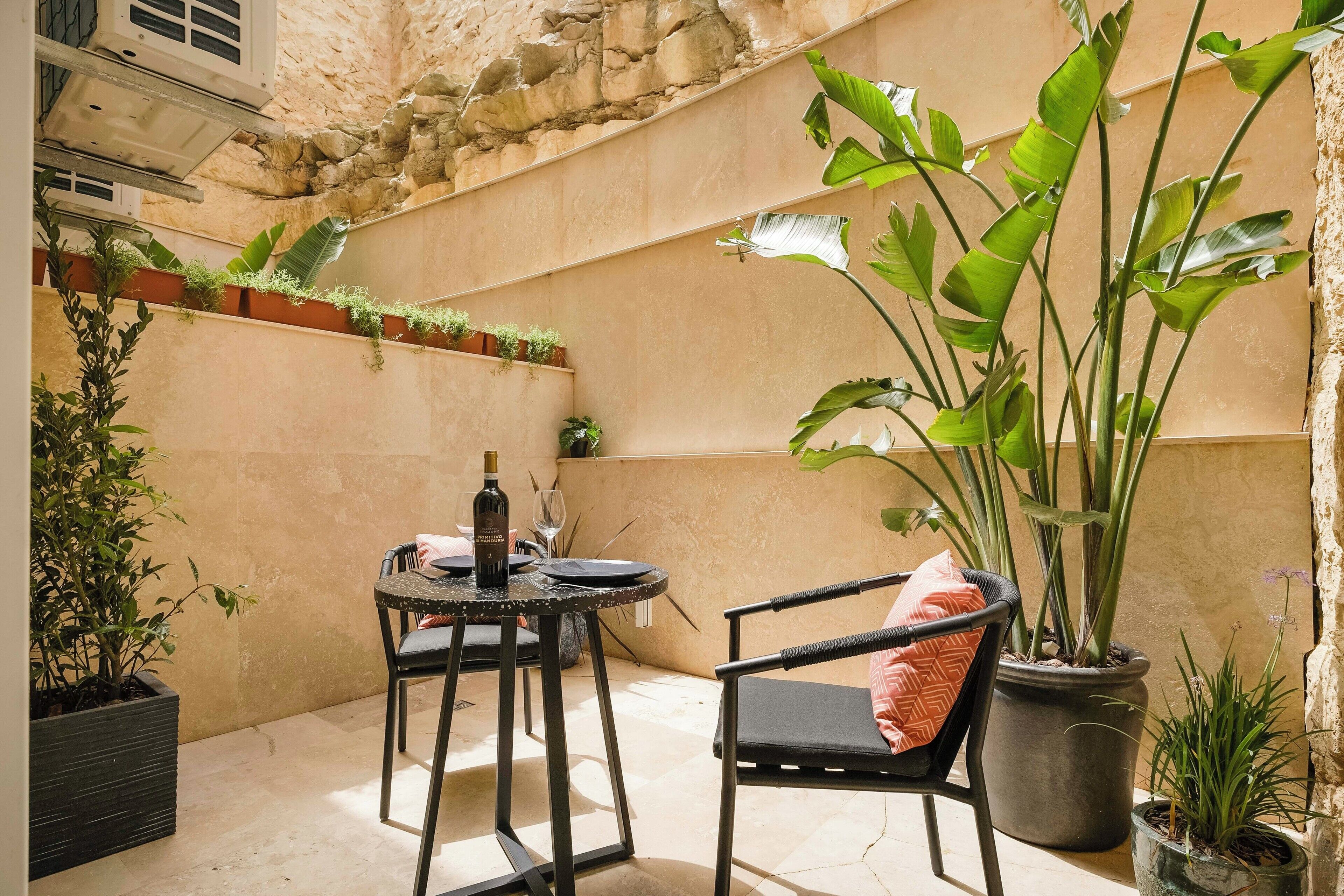 Um pátio externo com mesa, cadeiras, plantas em vasos e uma garrafa de vinho.
