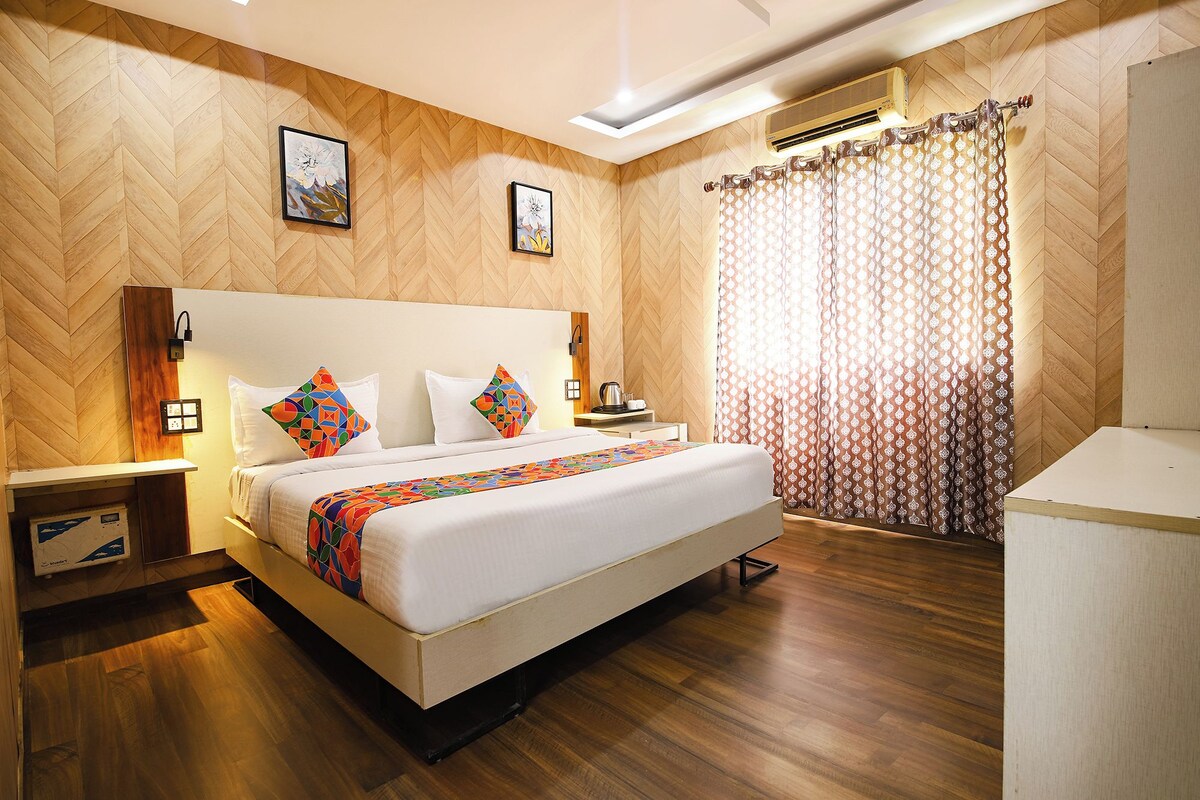 Quarto de hotel com uma cama grande, piso de madeira, cabeceira, duas mesinhas de cabeceira, um criado-mudo, ar condicionado fixado na parede e uma janela com cortinas estampadas.