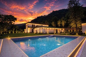 Piscina com borda azulejada, rodeada por área externa bem iluminada, com uma casa e árvores.