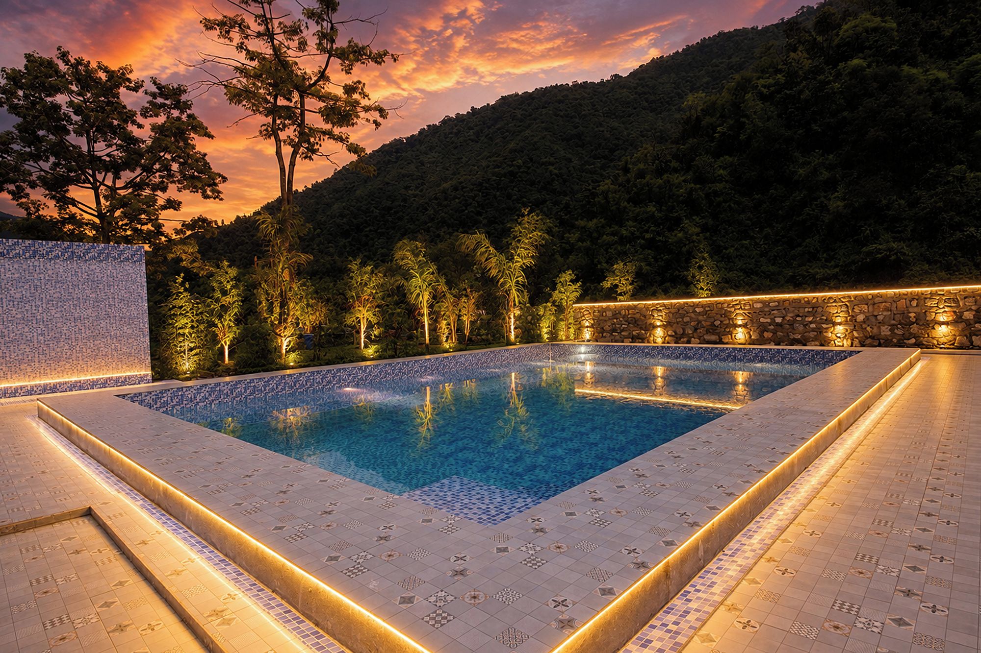 Uma piscina infinita retangular com borda azulejada, rodeada por um muro de pedra e uma fileira de árvores, com uma colina arborizada ao fundo durante o crepúsculo.