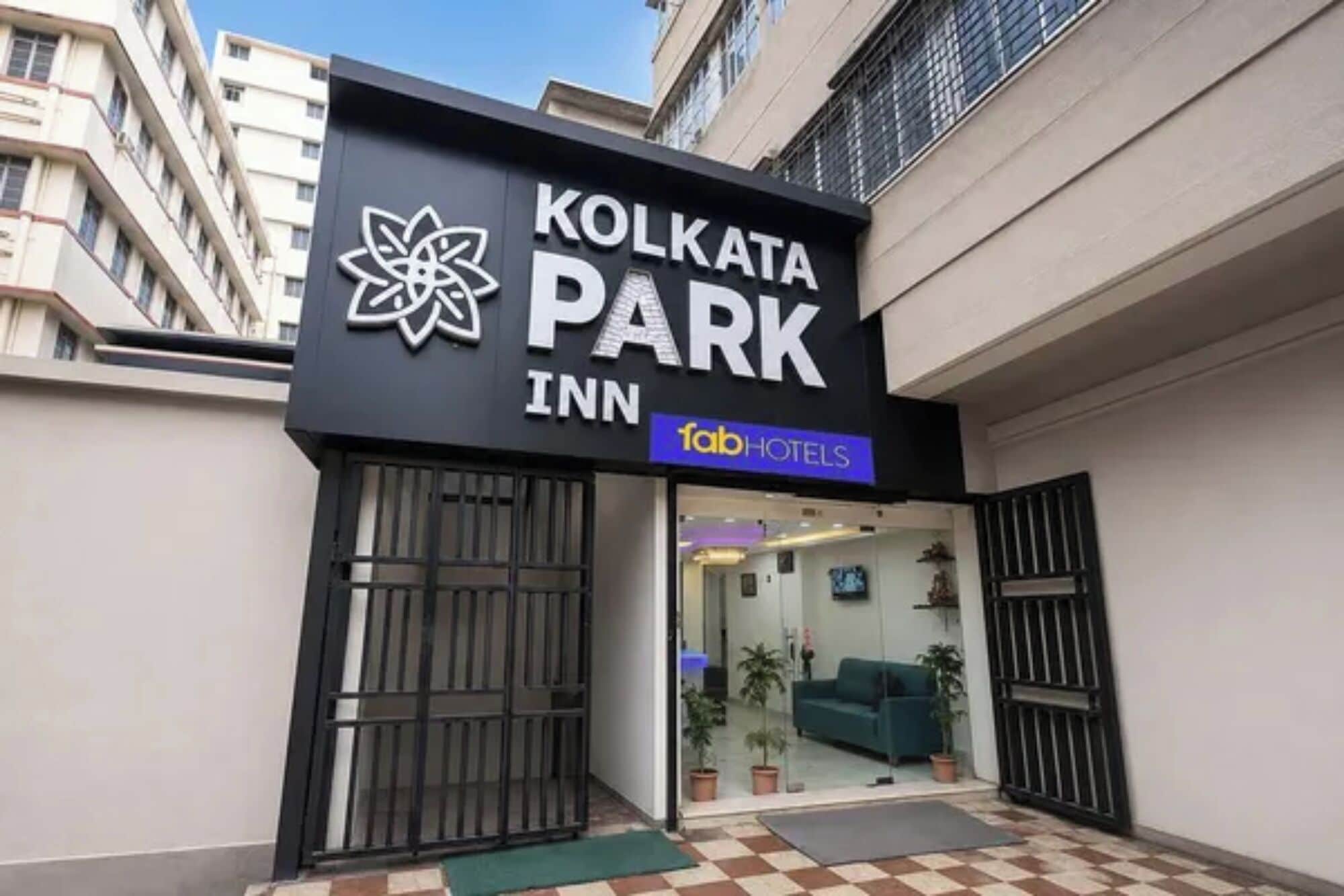 Exterior do Kolkata Park Inn, com fachada moderna, logotipo em destaque, entrada com portão e interior visível, com um sofá e plantas em vasos.