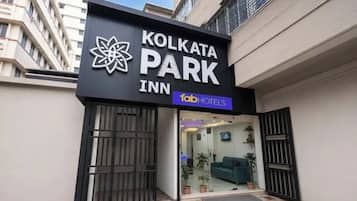Exterior do Kolkata Park Inn, com fachada moderna, logotipo em destaque, entrada com portão e interior visível, com um sofá e plantas em vasos.