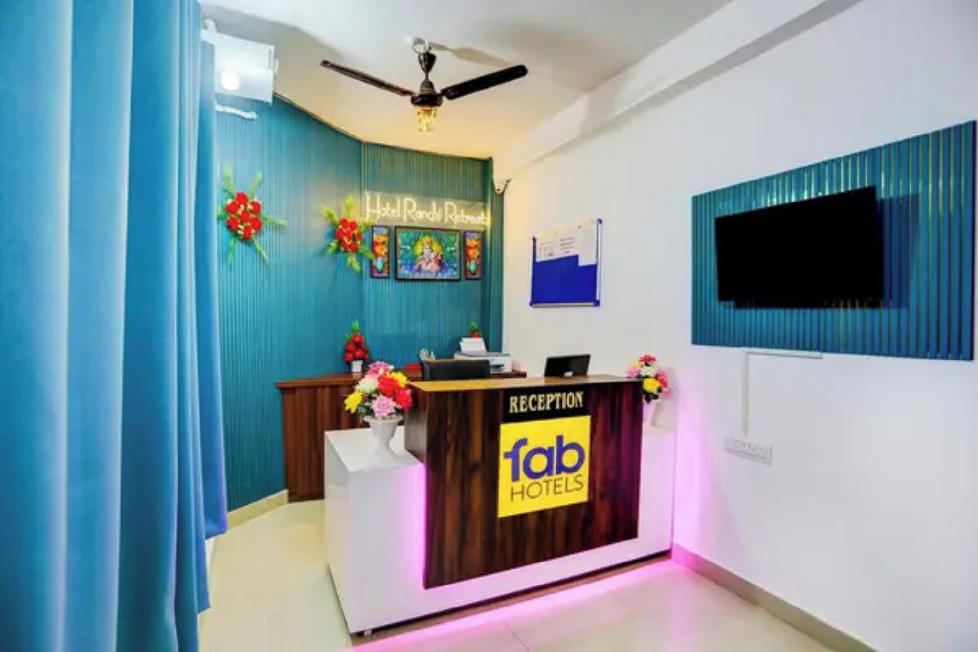 Uma mesa de recepção com uma placa indicando 'fab hotels', um ventilador de teto, uma televisão de tela plana fixada na parede e um mural de avisos.