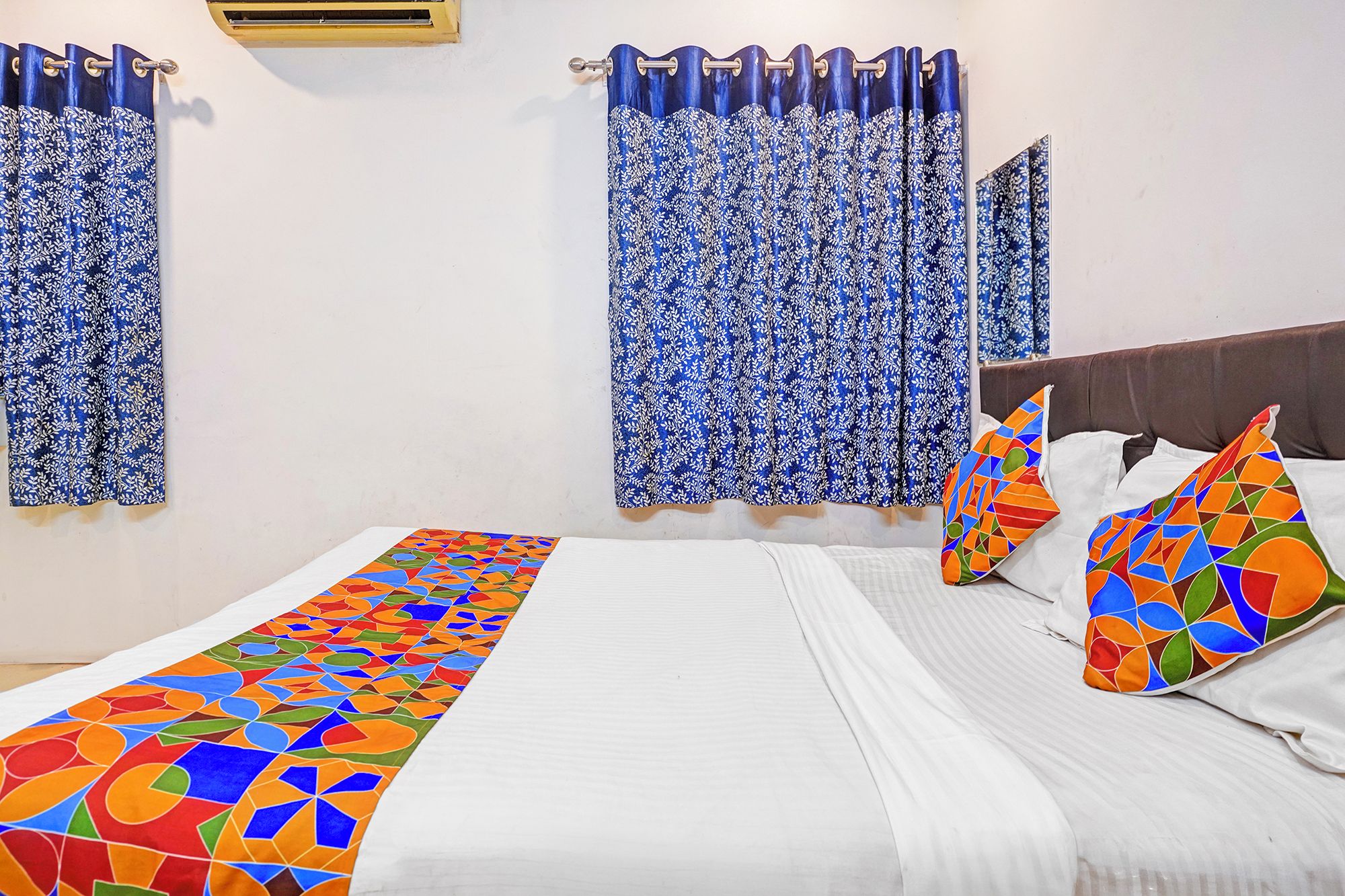Quarto de hotel com cama, roupa de cama branca, cobre-leito geométrico colorido e duas cortinas azuis estampadas, além de duas almofadas coloridas com padrão geométrico.