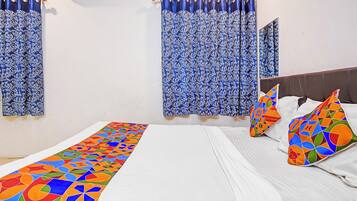 Quarto de hotel com cama, roupa de cama branca, cobre-leito geométrico colorido e duas cortinas azuis estampadas, além de duas almofadas coloridas com padrão geométrico.