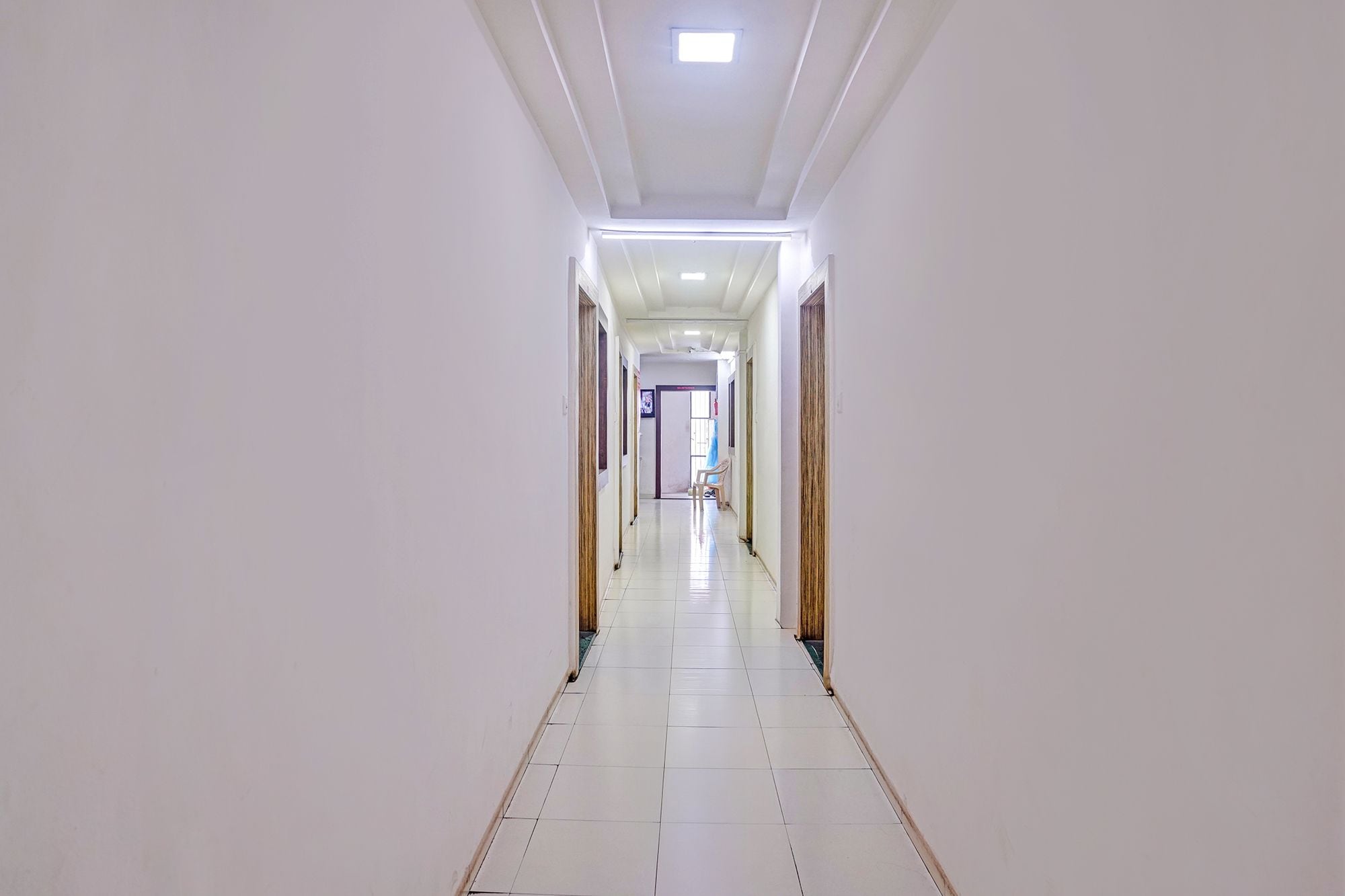 Um corredor comprido e estreito, com paredes brancas e chão de azulejos, que termina numa porta.