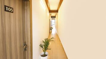 Corredor com porta de madeira com a placa '105', um vaso de planta e vista para um quarto no final.