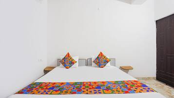 Uma cama com colcha de padrão geométrico colorido, duas almofadas com design similar, e uma porta de madeira escura à direita.