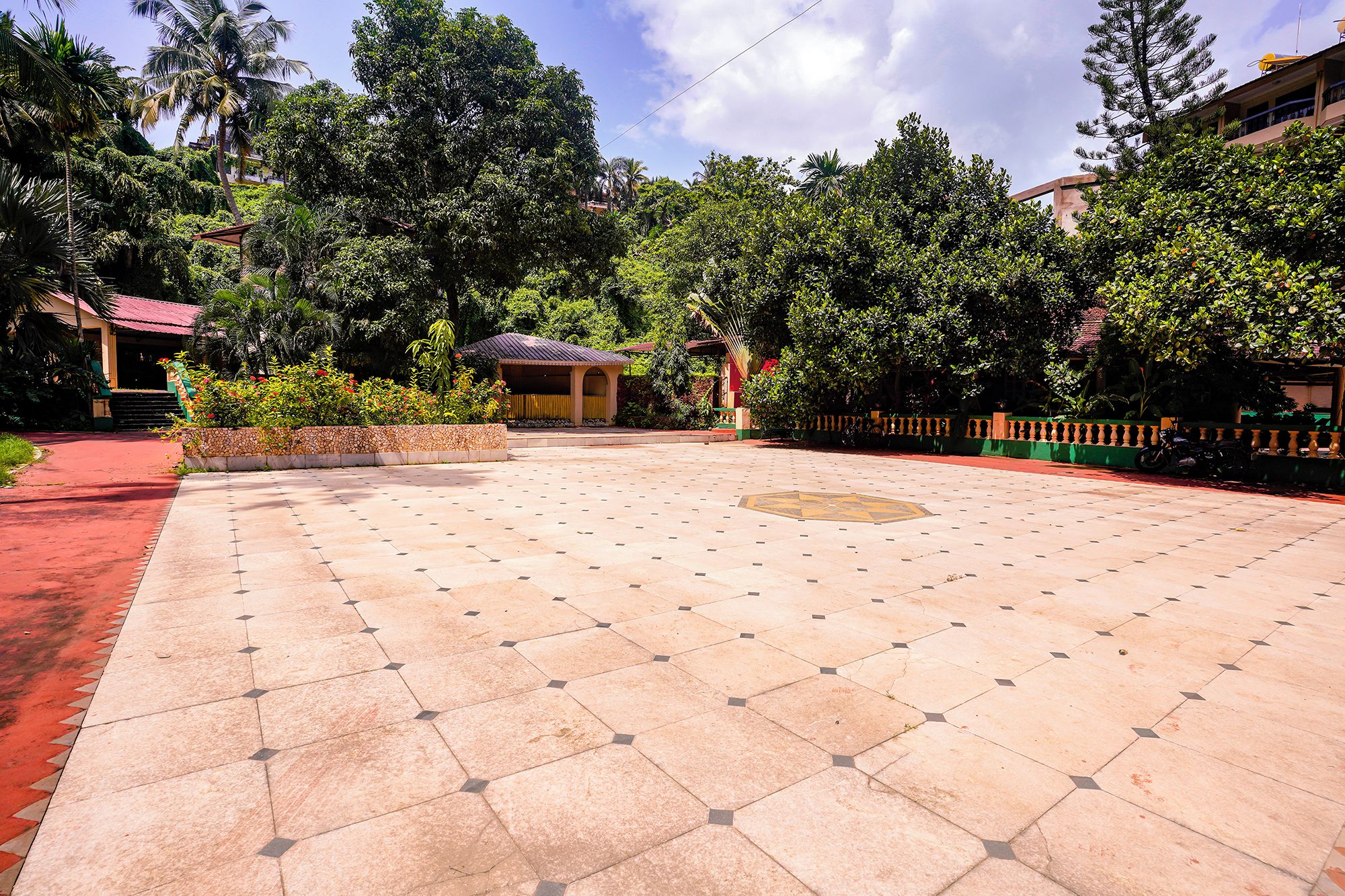Pátio com piso de azulejos, cercado por vegetação e um edifício com estrutura coberta.