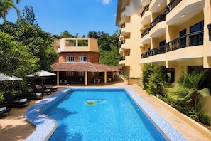Piscina com espreguiçadeiras e guarda-sóis, cercada por vegetação e um edifício com varandas.