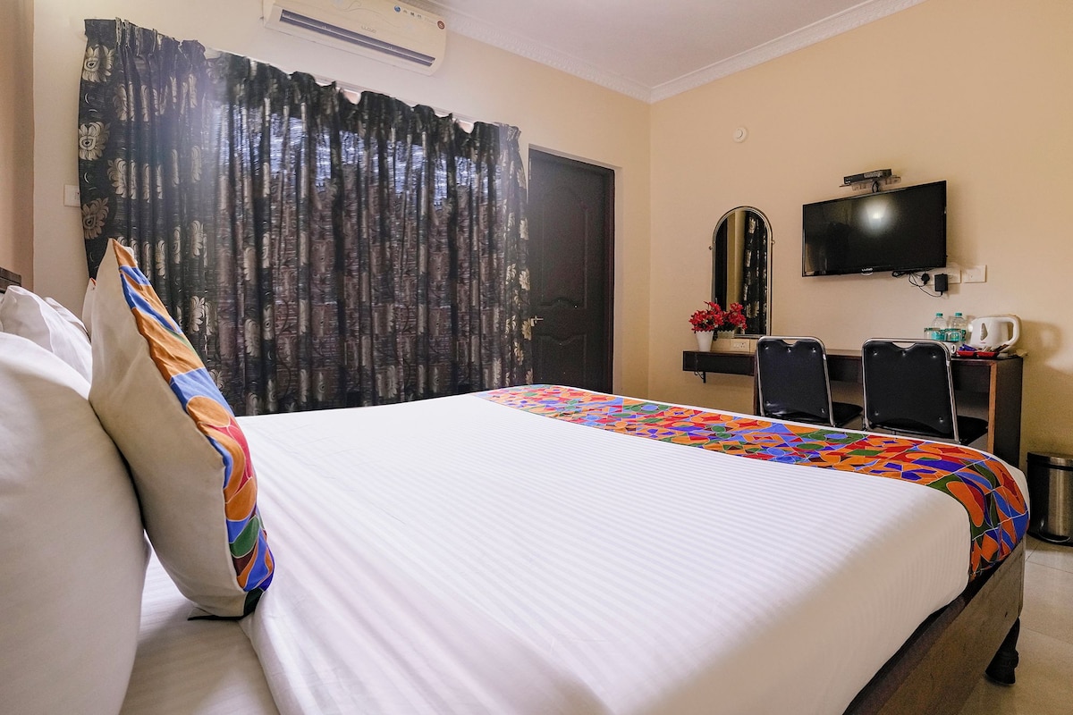 Quarto de hotel com cama, televisão, escrivaninha e cadeiras, e janela com cortinas.