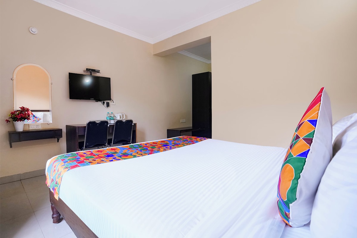 Quarto de hotel com cama, escrivaninha, cadeiras, televisão e espelho.