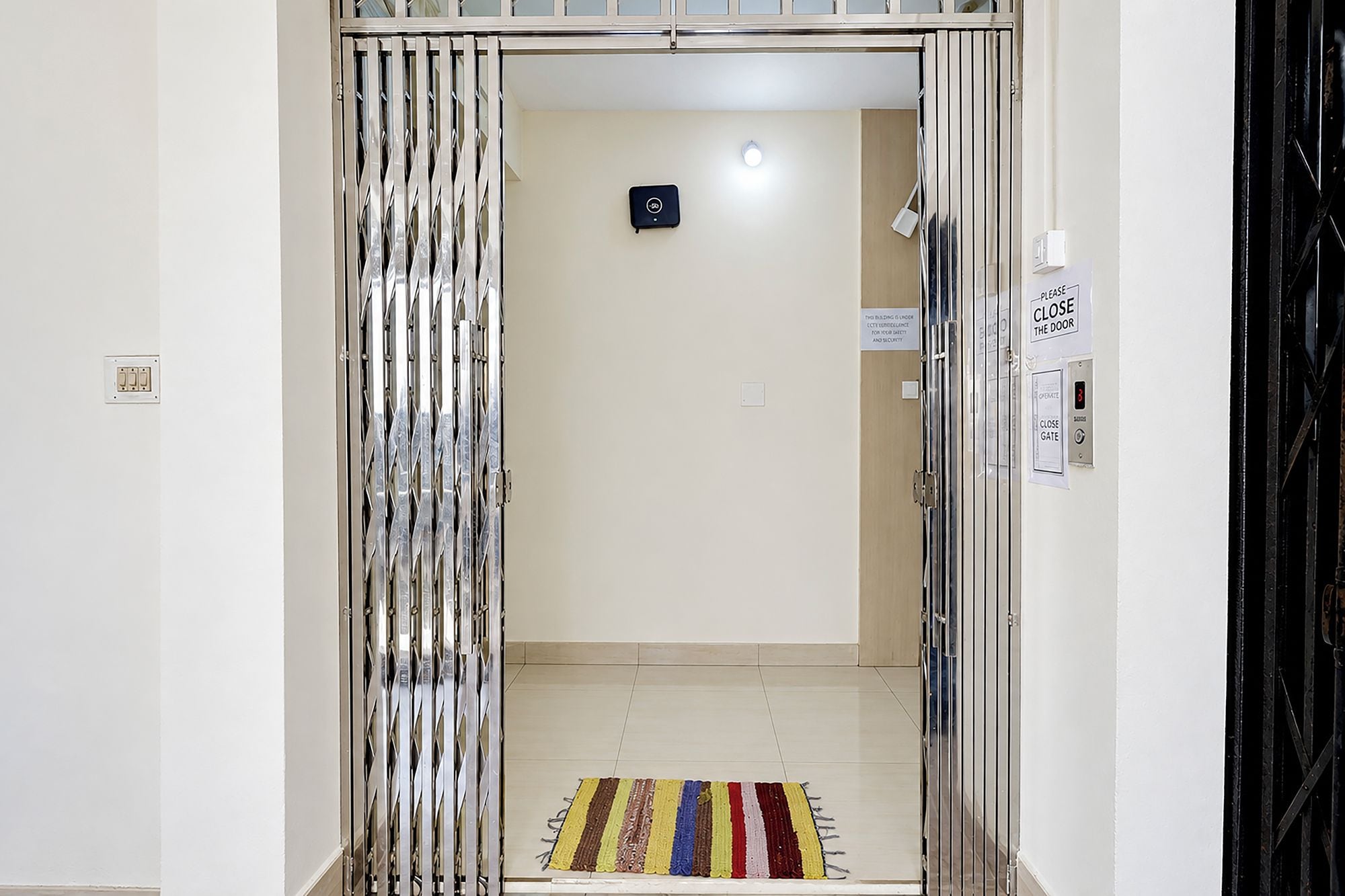Um elevador com um tapete listrado na base, uma placa indicando que o elevador está fora de serviço e uma porta com a mensagem 'Feche a Porta'.