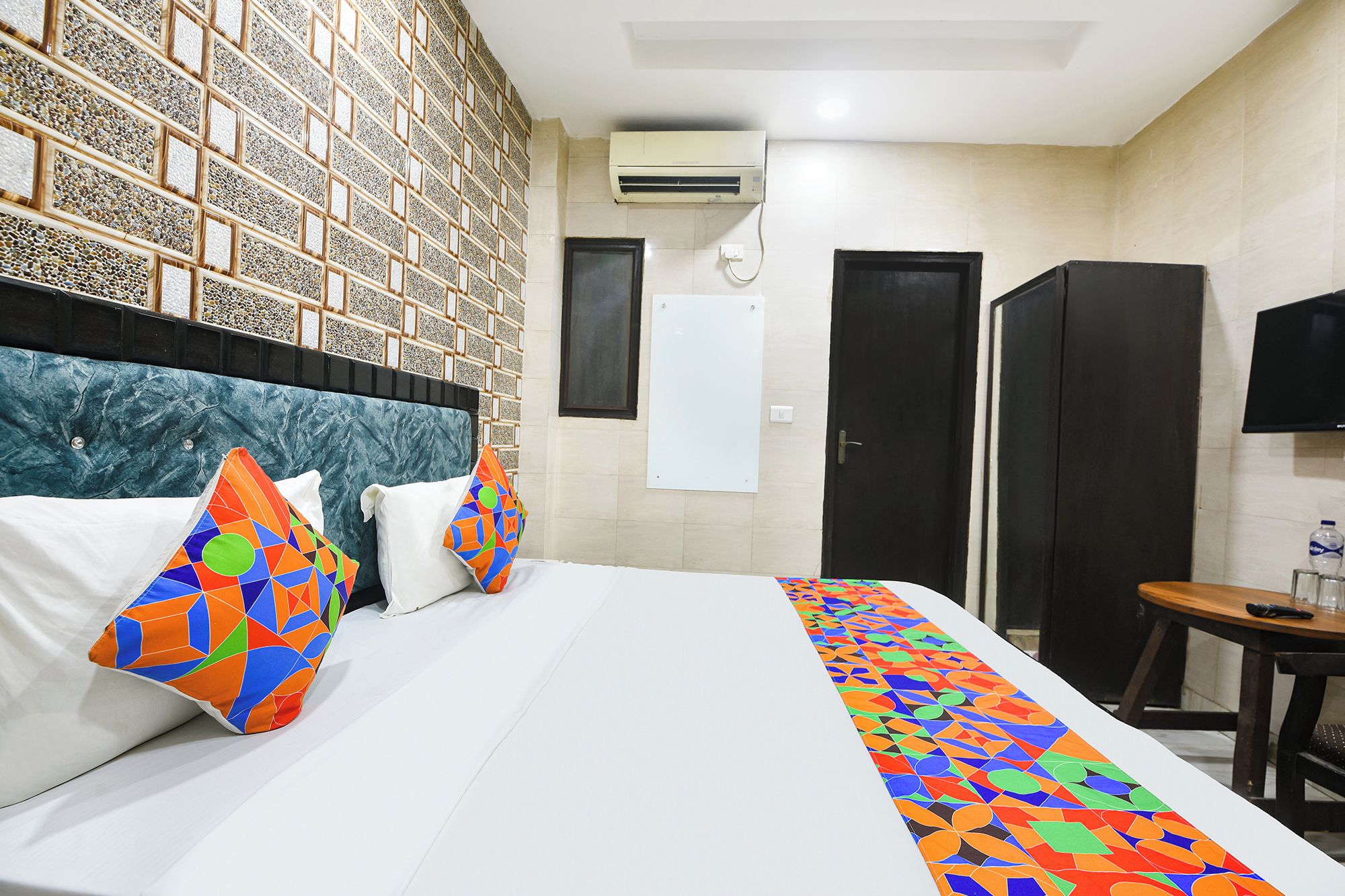 Quarto de hotel com parede estampada, cama com almofadas coloridas, televisão de tela plana e minibar.