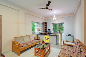 Quarto de hotel com cozinha compacta, dois sofás, uma mesa de centro de vidro e um ventilador de teto.