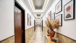 Corredor com obras de arte emolduradas nas paredes, porta de madeira e um banco com vaso e livros.