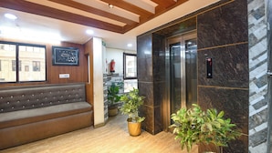 Um hall com sofá marrom, plantas em vasos, um elevador e uma parede de mármore.