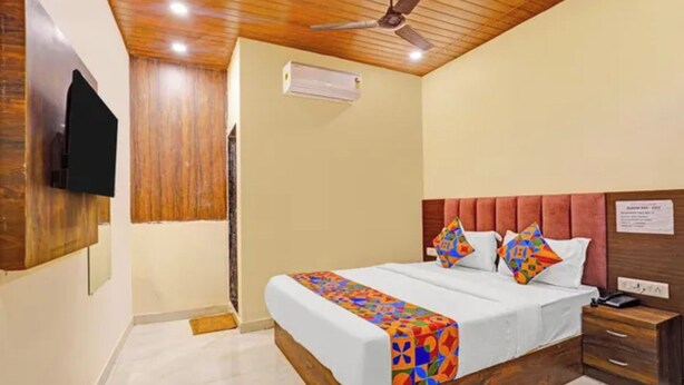 Quarto com teto de madeira, cama com roupa de cama colorida, mesa de cabeceira e televisão montada na parede.