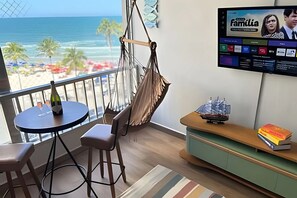 Uma varanda com vista para a praia, uma rede, uma mesa com uma garrafa e uma TV exibindo um programa.